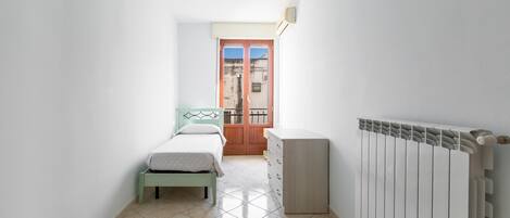 Quarto pequeno com uma cama, um radiador e uma porta para área externa.