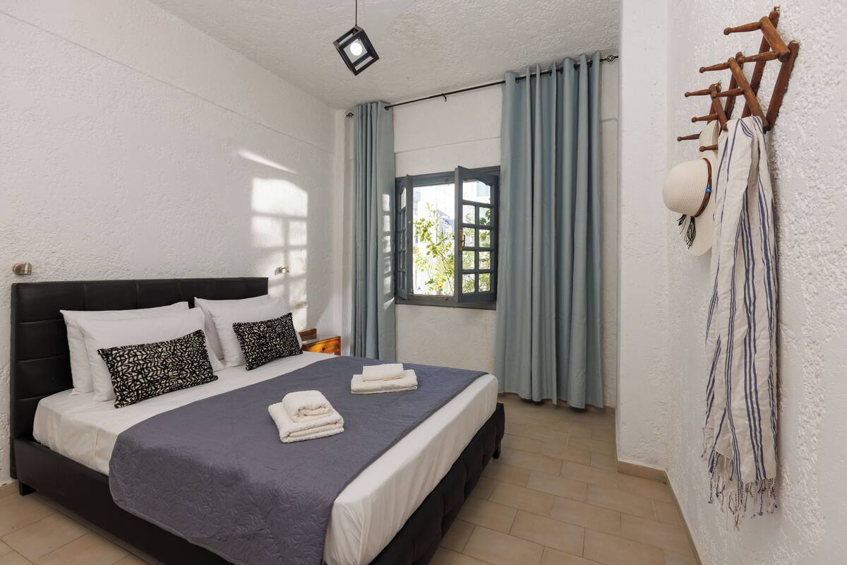 Cama bem arrumada com almofadas brancas e estampadas, edredom azul e janela com cortinas azul-claras.