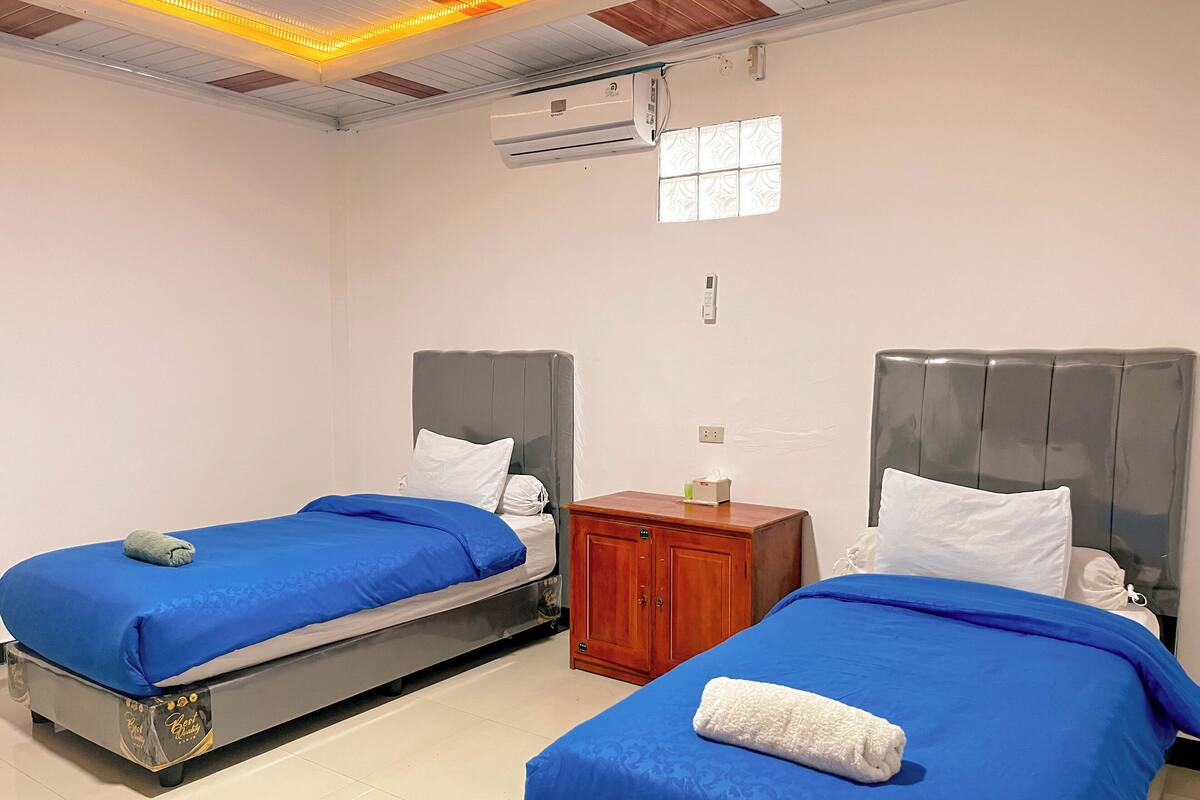 Quarto de hotel com duas camas, cada uma com colcha azul e almofadas brancas. Há uma mesa de cabeceira de madeira entre as camas com uma luminária pequena e um estojo de lenços. O quarto tem uma parede branca, uma janela com padrão decorativo e um aparelho de ar condicionado na parede.