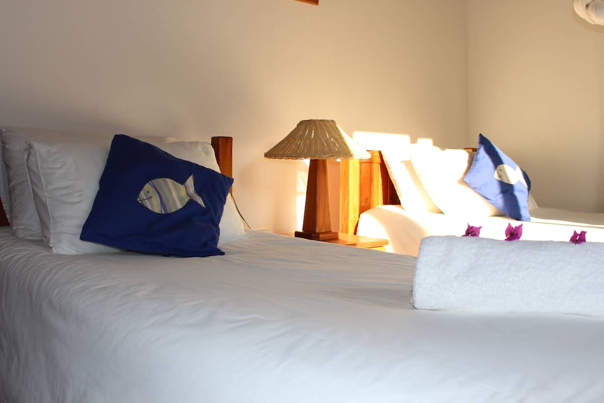 Cama bem arrumada com lençóis brancos e almofadas azuis, mesa de cabeceira de madeira com abajur, e janela com luz solar entrando.