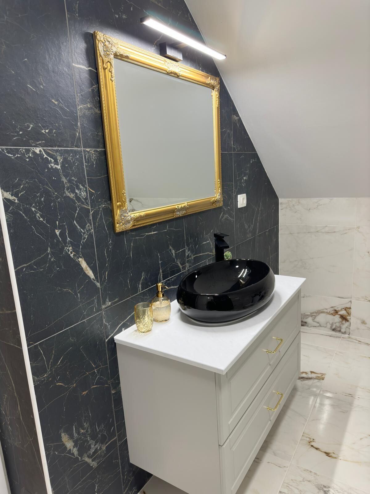 Um banheiro com parede de mármore preto, espelho com moldura dourada, pia preta e toucador branco com puxadores dourados.