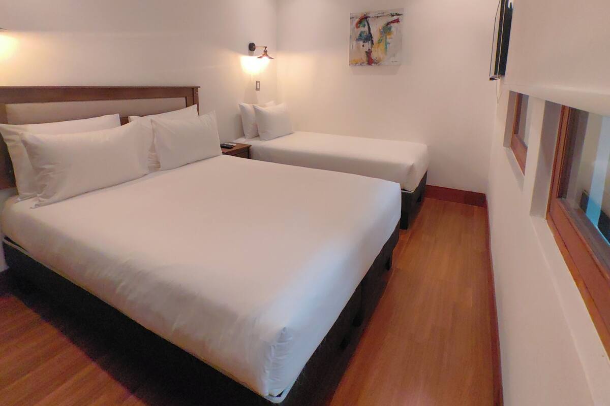 Quarto com uma cama grande, uma cama menor, piso de madeira, uma luminária no teto e um quadro na parede.