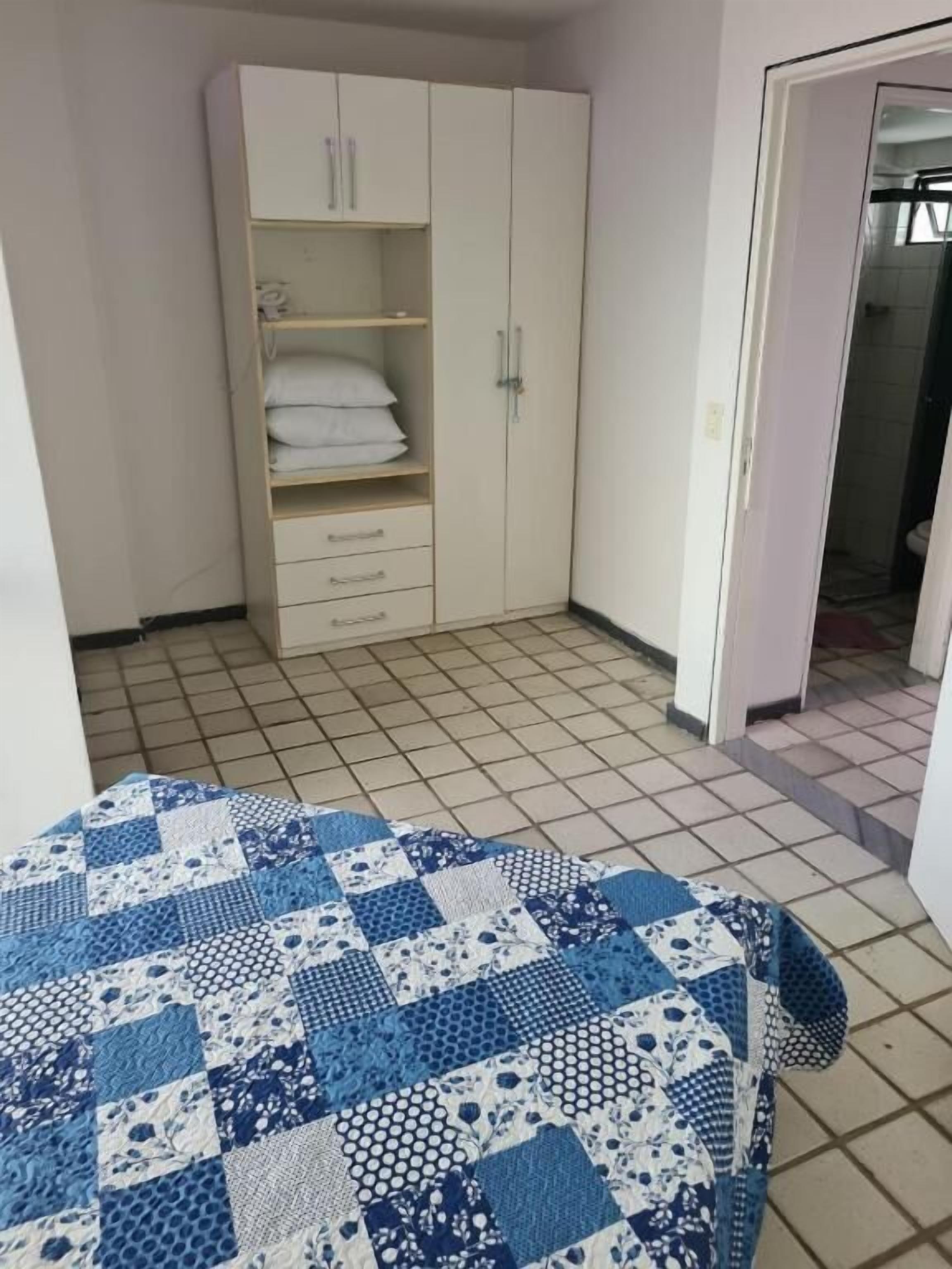 Quarto com colcha acolchoada, um guarda-roupa e um banheiro visível por uma porta aberta.