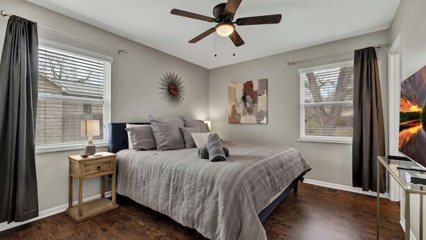 Um quarto com cama, mesa de cabeceira, ventilador de teto e duas janelas com cortinas.