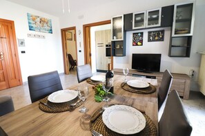 Uma sala de jantar com uma mesa de madeira posta para quatro pessoas, rodeada por cadeiras, e uma televisão fixada na parede.