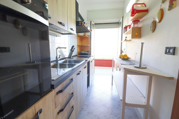 Cozinha com armários de madeira, forno preto e pia de aço inoxidável. Há uma mesa de madeira com um vaso e uma tigela de frutas no balcão.