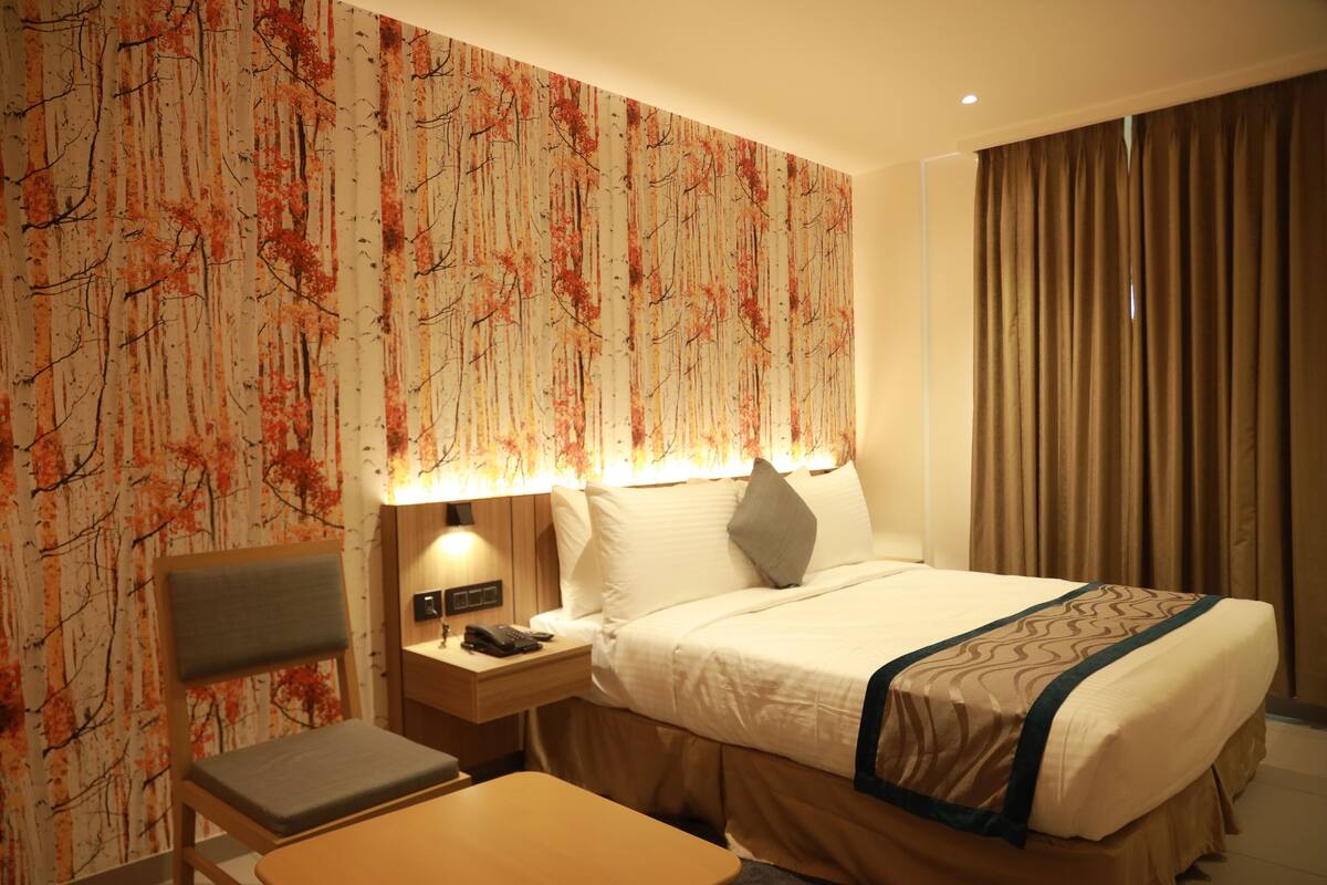 Quarto de hotel com cama, mesa de cabeceira com abajur, cadeira e janela com cortinas.