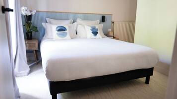 Cama bem arrumada com roupa de cama branca e almofadas decorativas, mesa de cabeceira com abajur e uma planta em vaso.