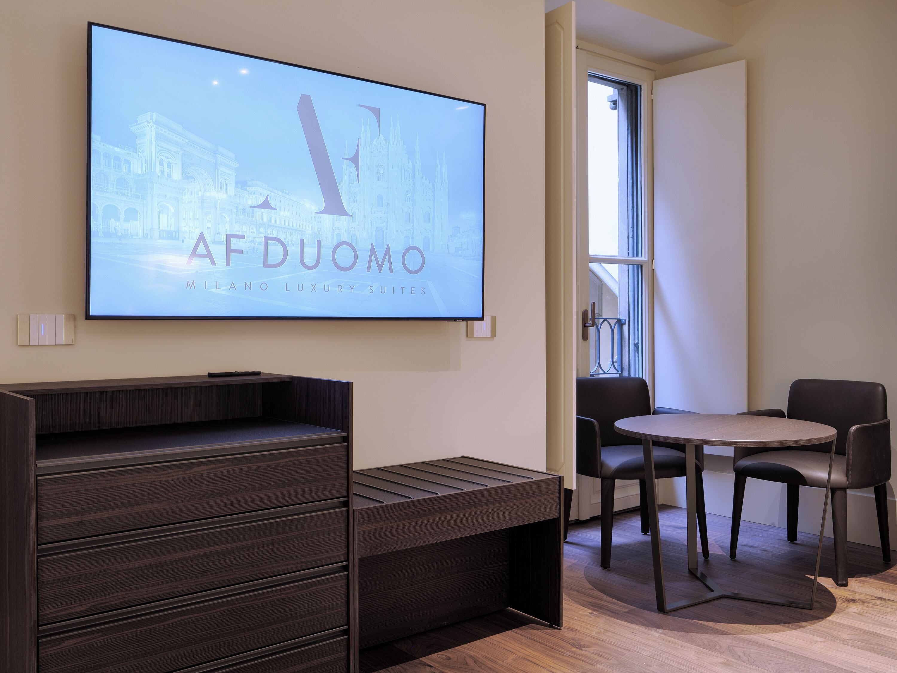 Quarto de hotel moderno com uma grande tela de TV exibindo 'AF DUOMO MILANO LUXURY SUITES'. Há uma mesa consola de madeira escura, uma mesa redonda com quatro cadeiras e uma janela com vista para a cidade.