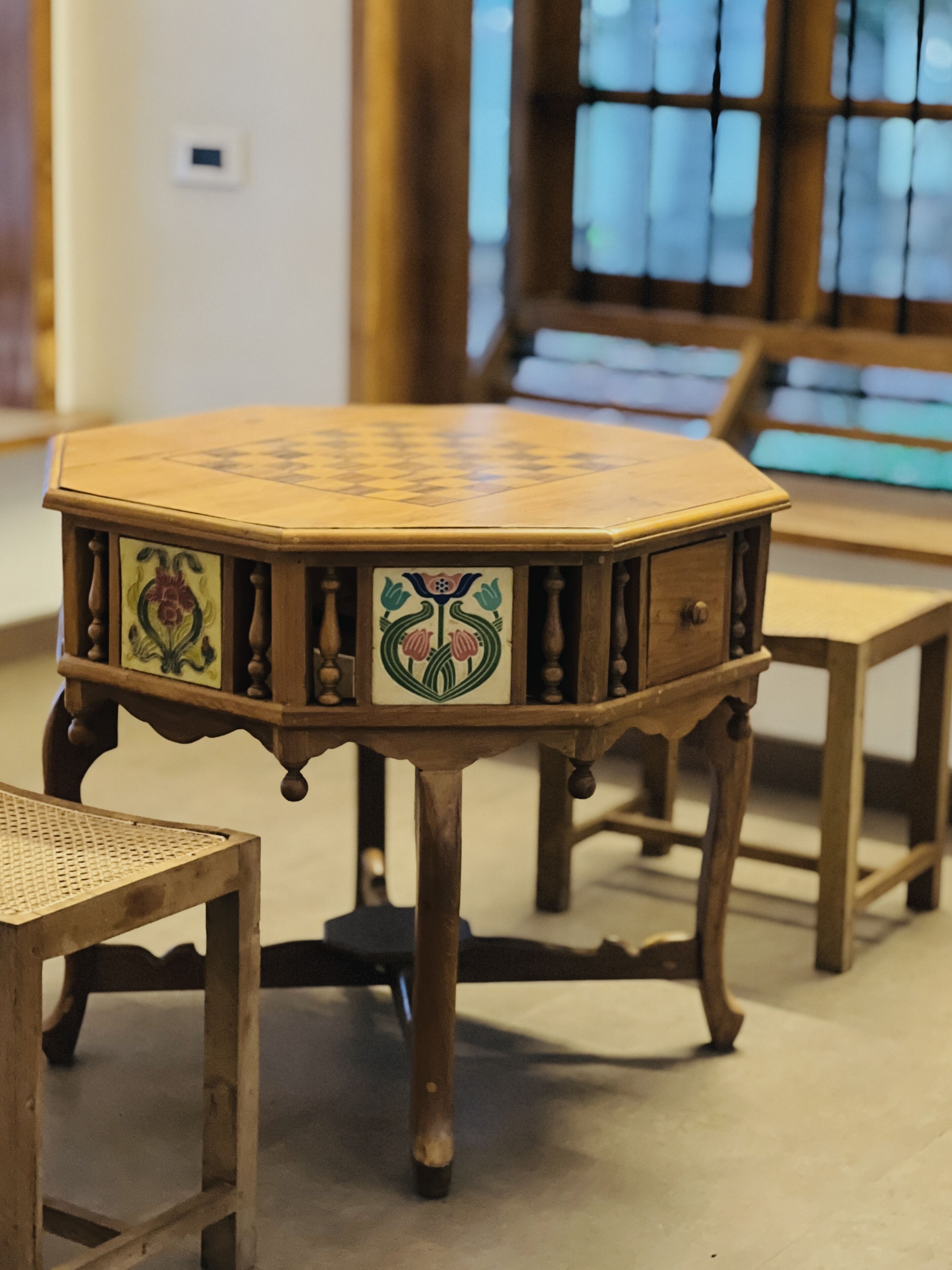 Uma mesa de madeira com desenhos elaborados, uma cadeira combinando e uma janela com padrão de grade ao fundo.