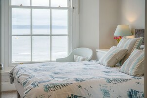 Quarto com uma grande janela com vista para o mar, cama com roupa de cama listrada, uma cadeira e mesa de cabeceira com abajur e flores.