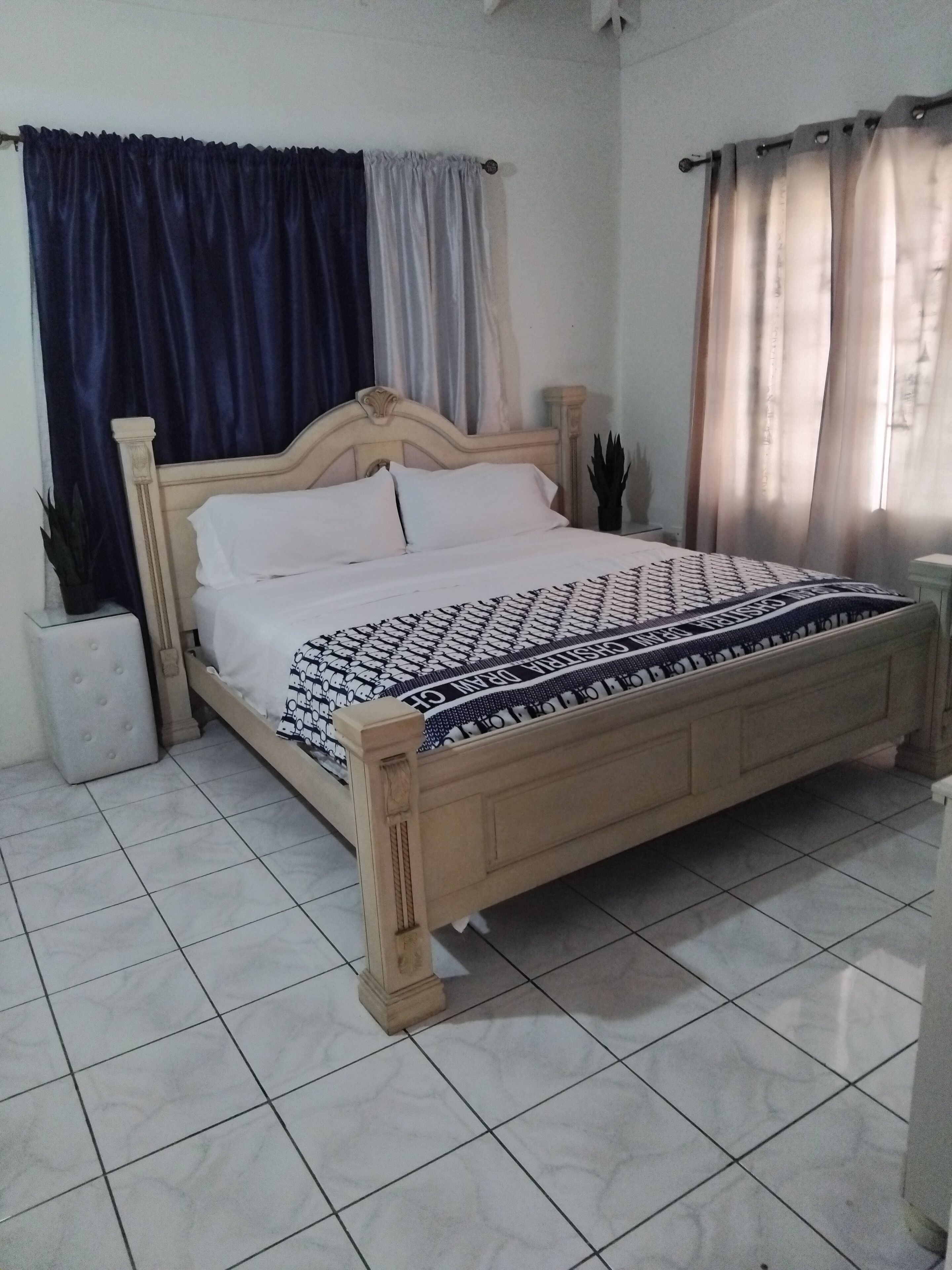 Uma cama de solteiro com colcha estampada, uma mesinha de cabeceira com planta em vaso e uma janela com cortinas.