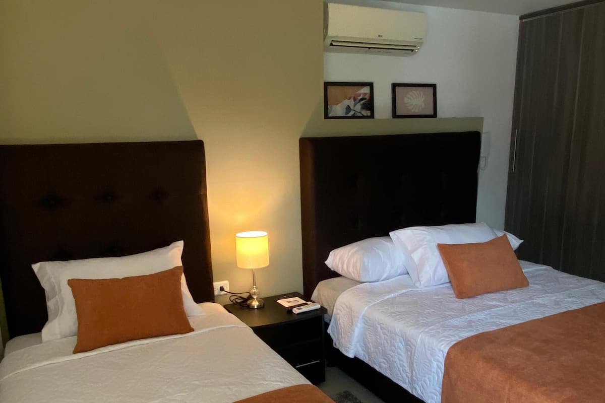Quarto de hotel com duas camas, uma mesa de cabeceira com abajur e um ventilador de teto.