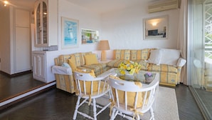 Uma sala de estar com sofá, cadeiras, mesa de centro de vidro e um armário.