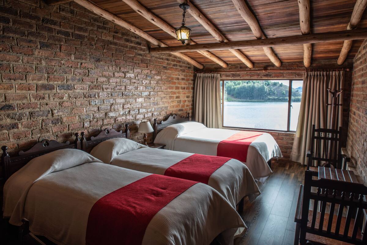 Quarto com duas camas, teto de madeira, parede de tijolos, janela com vista para um lago e um banco.