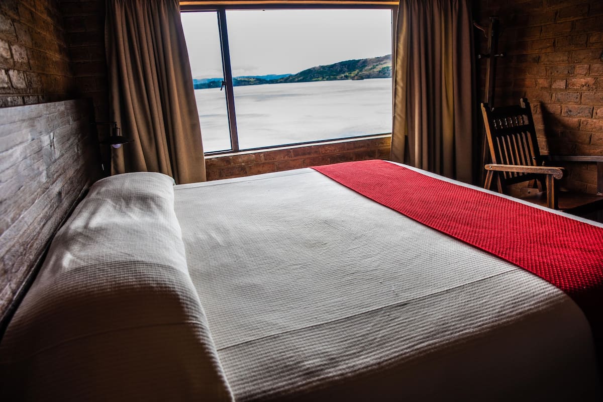 Uma cama com um tapete vermelho, uma cadeira de madeira e vista para um corpo d'água através de uma janela com cortinas.