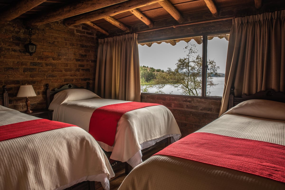 Quarto com três camas, parede de tijolos, janela com cortinas e uma luminária.