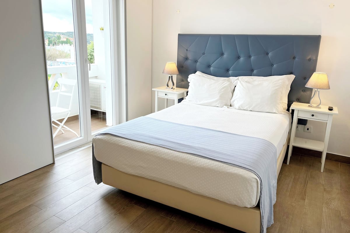 Quarto bem arrumado com uma cama grande, duas mesas de cabeceira com luminárias e uma porta de vidro deslizante para uma área externa.