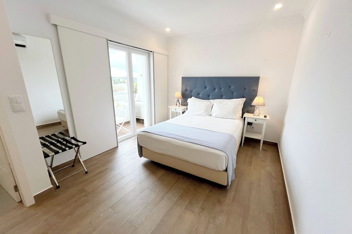 Quarto moderno com uma cama grande, uma porta de vidro deslizante para uma varanda e uma pequena mesa com abajur.