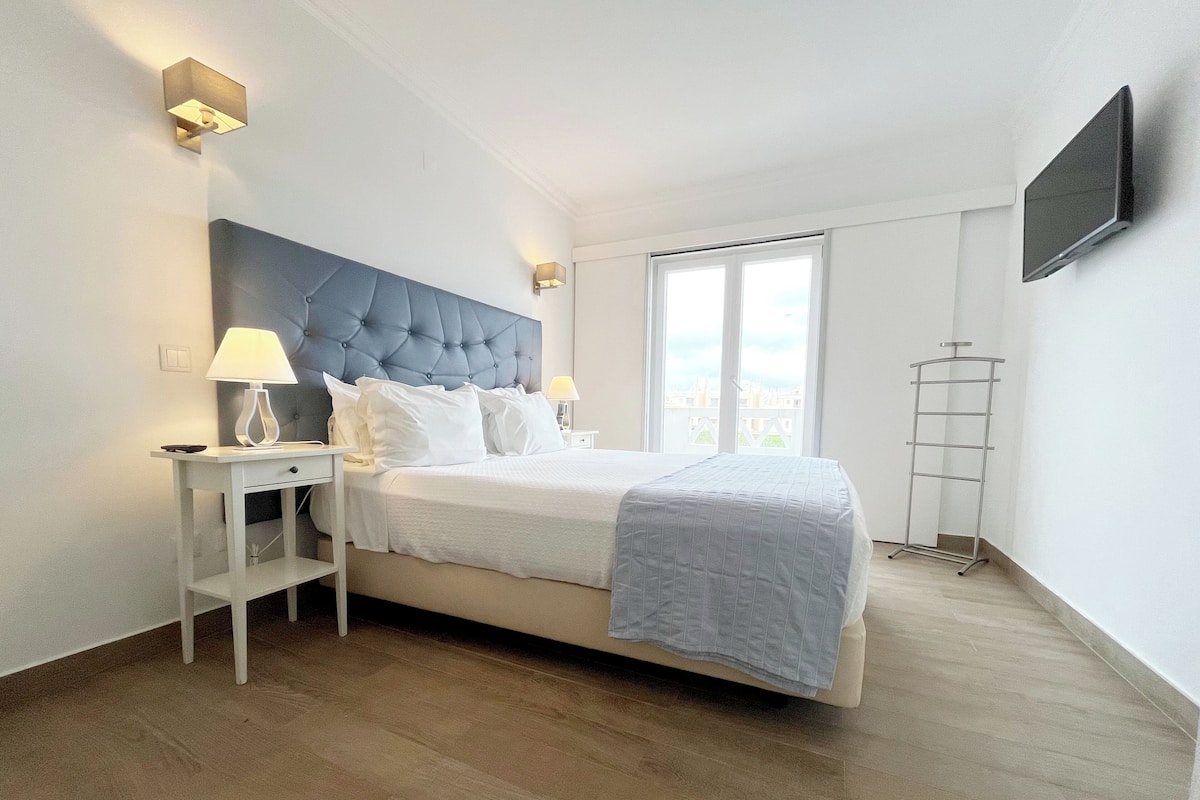 Quarto bem arrumado com uma cama grande, luminárias de cabeceira, mesa de cabeceira, televisão montada na parede e uma janela com vista.