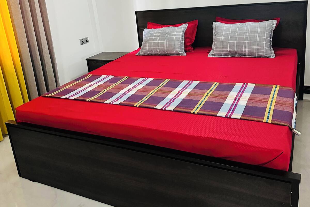 Uma cama com colcha vermelha e duas almofadas, cabeceira preta e luminária sobre a cama.
