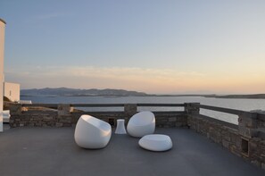 Terraço no telhado com assentos brancos modernos e balaustrada de pedra, com vista para um corpo d'água e colinas ao longe.