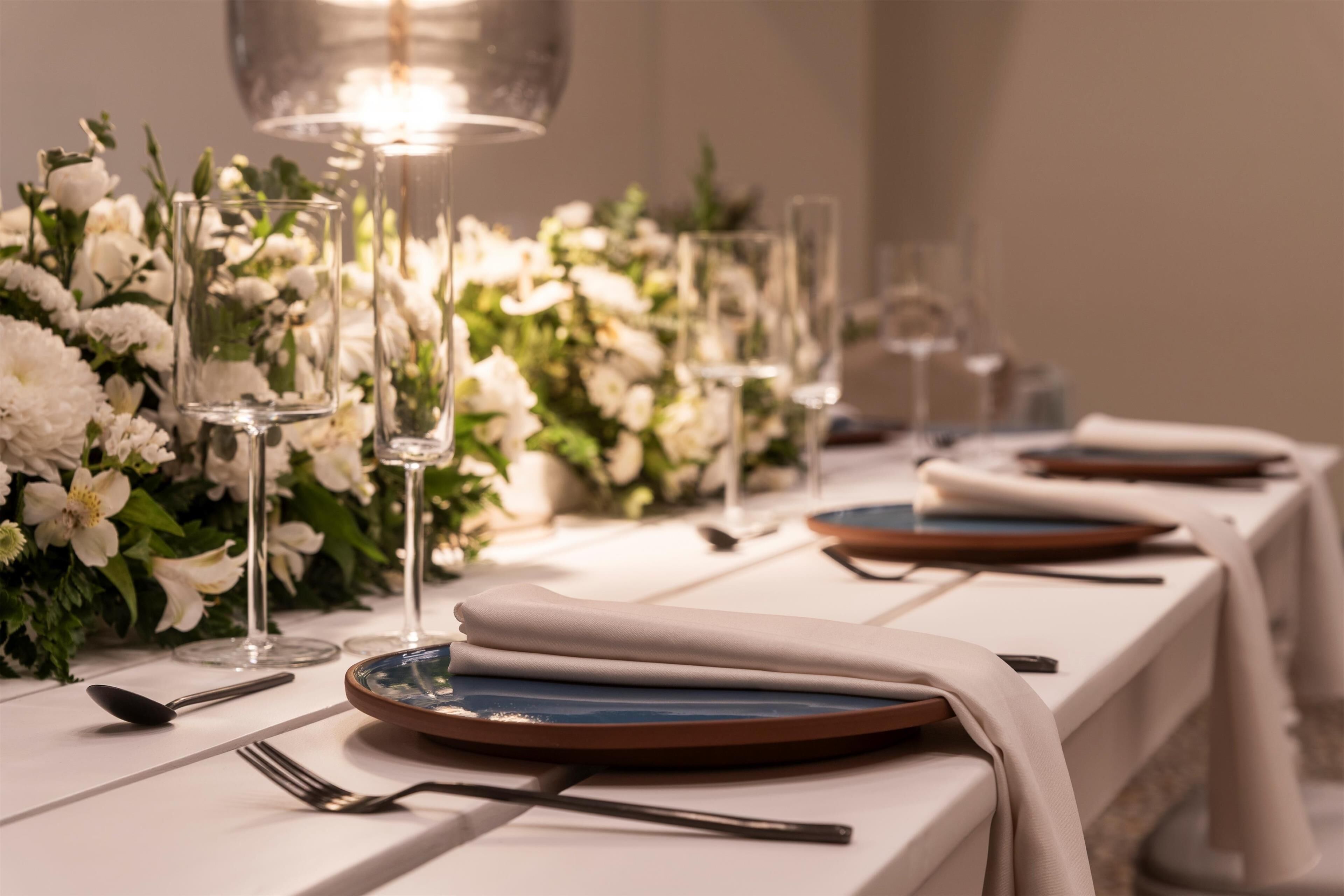 Uma mesa posta com pratos, copos e talheres, rodeada por flores brancas e folhagem.