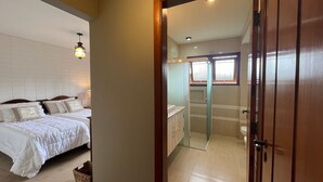 Quarto com duas camas, banheiro com chuveiro de vidro e corredor que leva a uma porta.