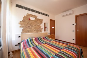 Quarto com edredom listrado e colorido, decoração de parede em pedra, cabeceira de madeira, luminária de mesa e porta de madeira.