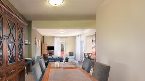 Uma sala de jantar com uma mesa e cadeiras de madeira, um armário com portas de vidro e uma televisão ao fundo.