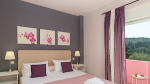 Quarto com cama, duas mesas de cabeceira, janela com cortinas e três quadros de orquídeas na parede.