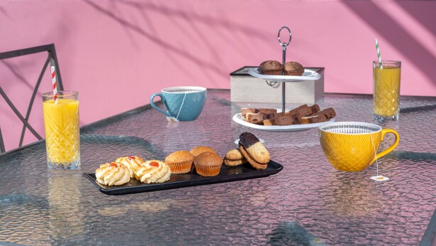Uma mesa com uma bandeja de muffins, um bule, uma xícara, um copo de suco de laranja e uma caixa de biscoitos.