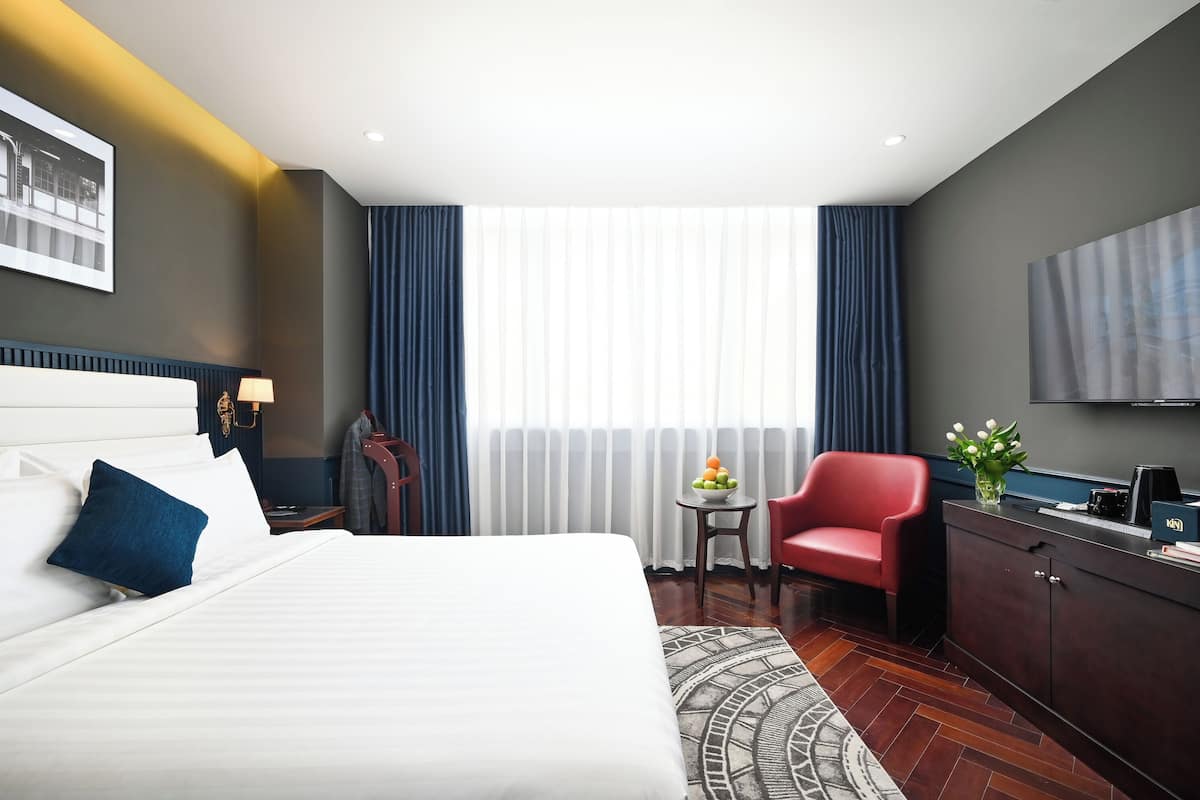 Quarto de hotel com cama, escrivaninha, cadeira, mesa pequena com uma fruteira, um vaso com flores e TV na parede.