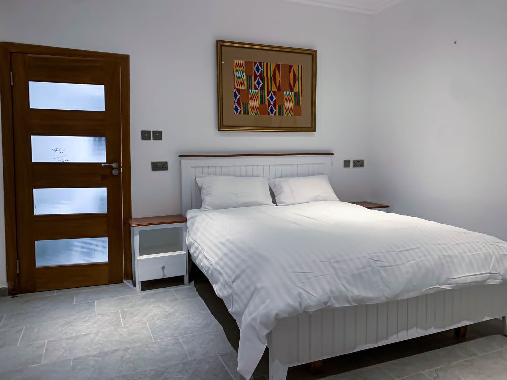 Quarto com cama, mesa de cabeceira, gaveta, porta de madeira e uma obra de arte emoldurada na parede.