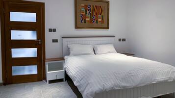 Quarto com cama, mesa de cabeceira, gaveta, porta de madeira e uma obra de arte emoldurada na parede.