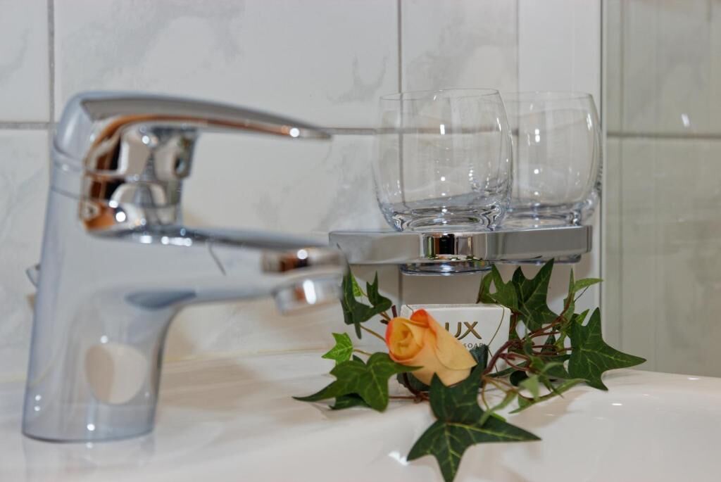 Uma casa de banho com uma torneira moderna, dois copos de vidro transparentes numa bandeja e um pequeno vaso com uma rosa amarela e folhas verdes.