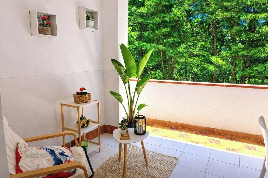 Uma varanda com vista para árvores, uma mesinha com uma planta, uma cadeira e uma estante com objetos decorativos.