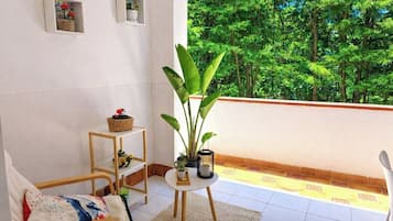 Uma varanda com vista para árvores, uma mesinha com uma planta, uma cadeira e uma estante com objetos decorativos.