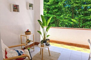Uma varanda com vista para árvores, uma mesinha com uma planta, uma cadeira e uma estante com objetos decorativos.