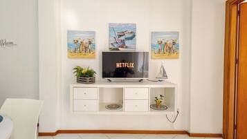 Uma sala de estar com um rack branco, uma TV exibindo 'NETFLIX' e três quadros com tema praiano acima.