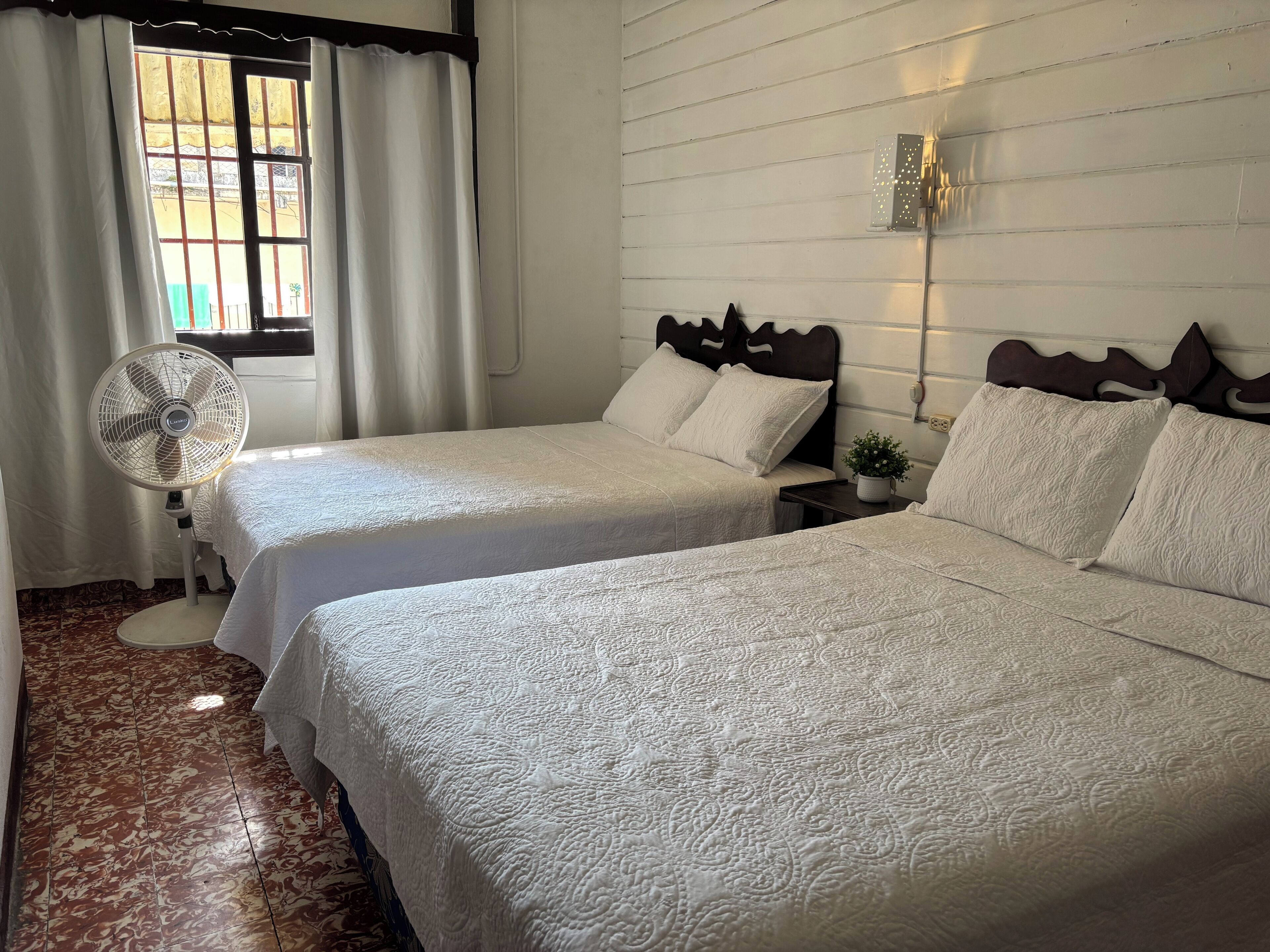 Quarto de hotel com duas camas, um ventilador, uma mesinha com uma planta e uma janela com cortinas.