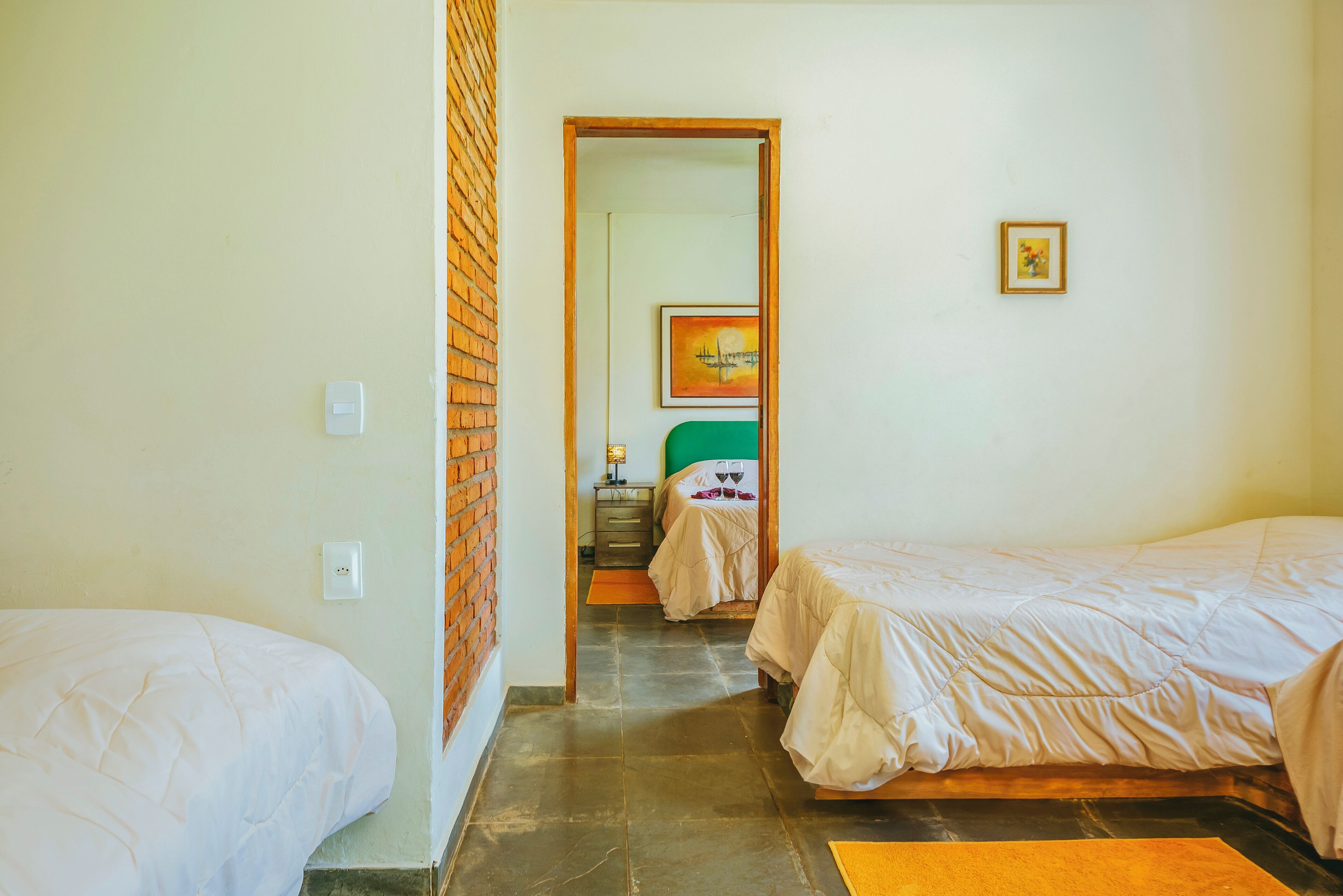 Quarto com duas camas, parede de tijolos, porta para outro cômodo e um quadro na parede.