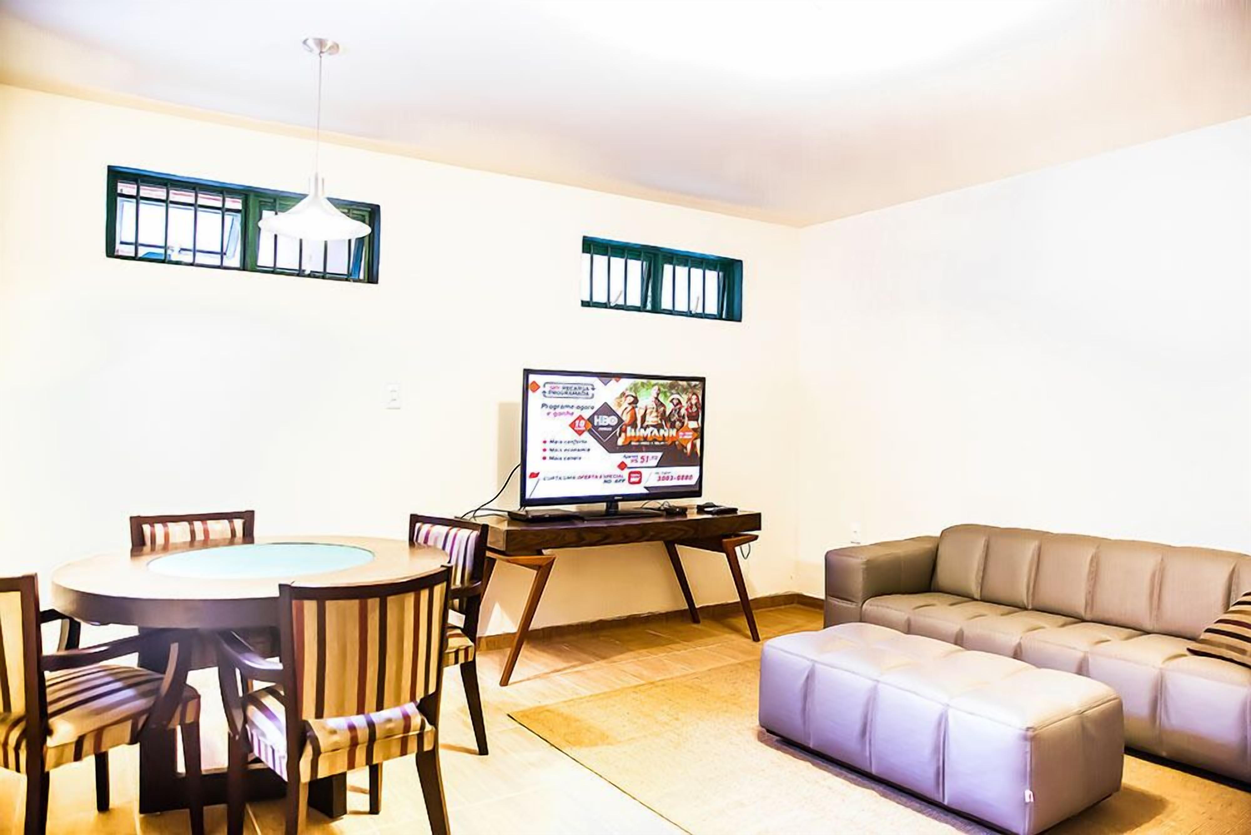 Sala de estar com mesa de jantar redonda, cadeiras, sofá e televisão exibindo um programa.