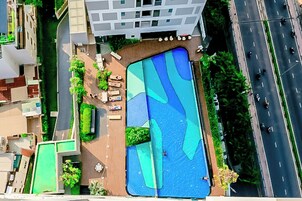 Vista aérea de um edifício moderno com piscina no terraço, rodeado por áreas verdes e uma estrada.
