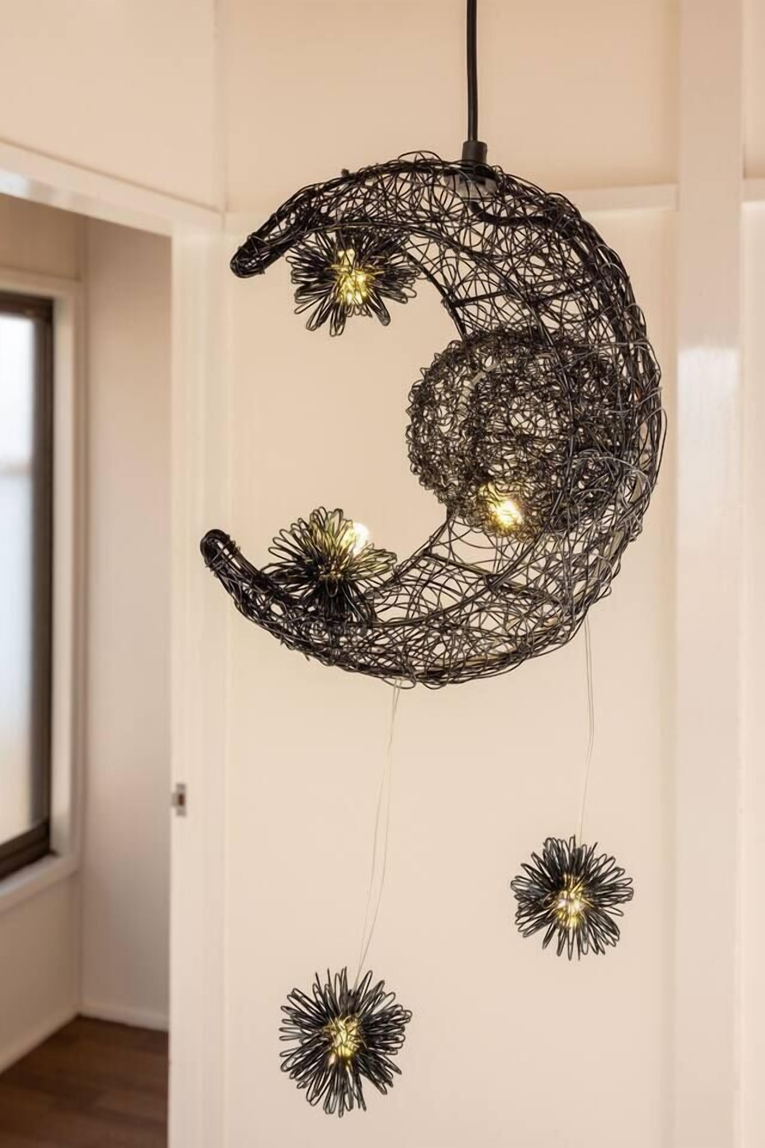 Um candeeiro em forma de lua com estrutura de arame pendurado, com três flores menores também de arame abaixo.