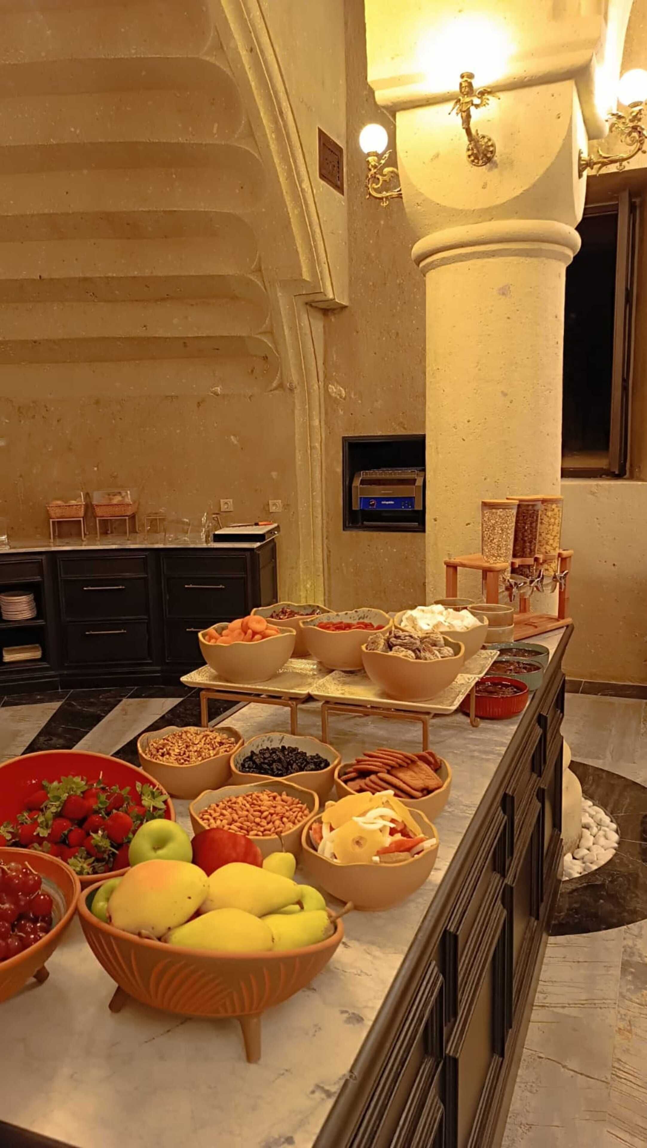 Disposição de buffet com diversos alimentos em tigelas e recipientes, piso de mármore e uma coluna grande ao fundo.