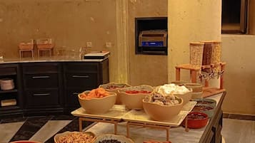 Disposição de buffet com diversos alimentos em tigelas e recipientes, piso de mármore e uma coluna grande ao fundo.