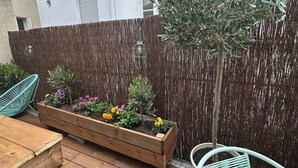 Um deck de madeira com um canteiro embutido, contendo diversas plantas e flores. Há duas cadeiras de metal verde e uma mesa de madeira redonda. Uma árvore alta é visível ao fundo.