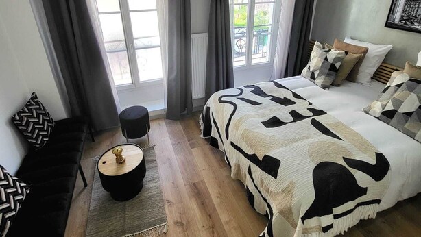 Um quarto moderno com uma cama grande, uma mesinha redonda, um sofá e vista para o exterior por grandes janelas.