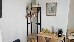 Um espaço compacto com um sofá azul, uma pequena mesa redonda com um vaso, uma escrivaninha de madeira com uma cadeira, uma estante de metal preta com cestos e um mapa emoldurado na parede.